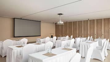 Sala de conferências com várias mesas, cadeiras, tela de projetor e parede de painéis de madeira.
