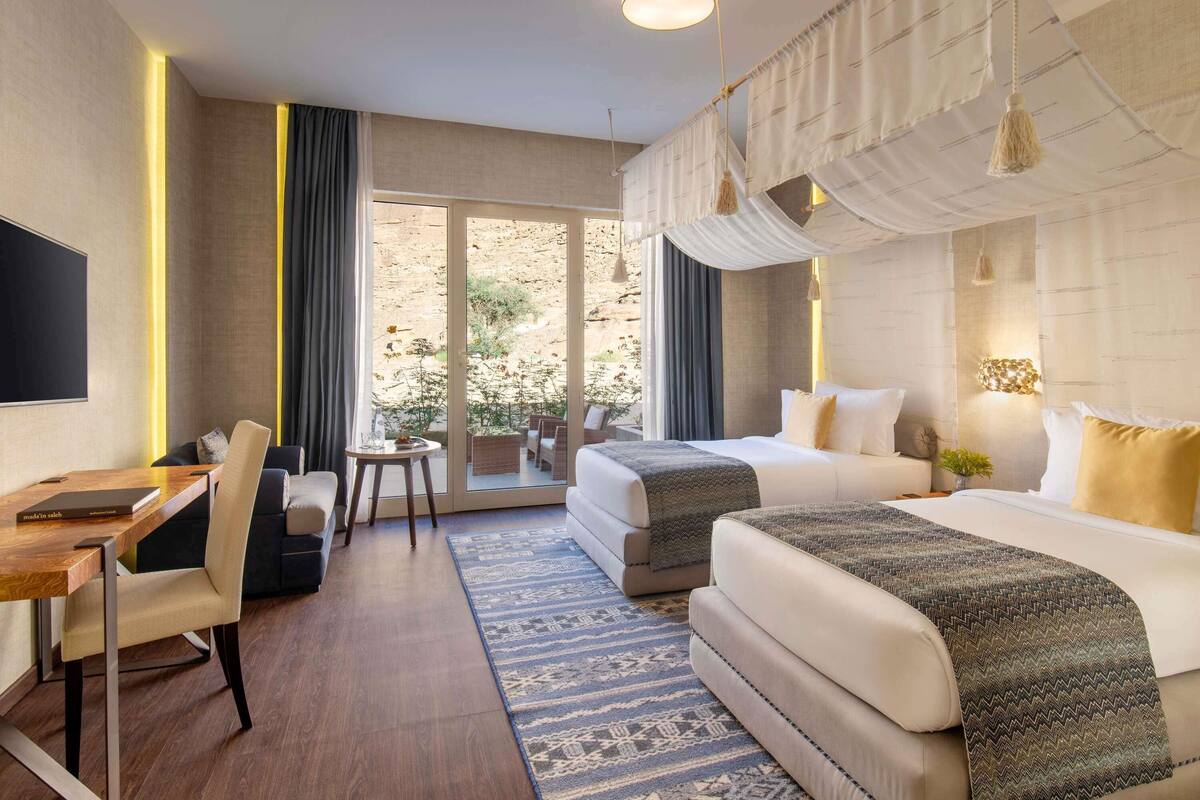 Quarto de hotel com secretária, duas camas, televisão e uma janela grande com cortinas.