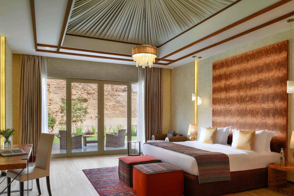 Quarto de hotel com uma cama grande, uma escrivaninha, uma cadeira, um sofá, um banquinho vermelho e vista para uma paisagem rochosa através da porta de vidro.