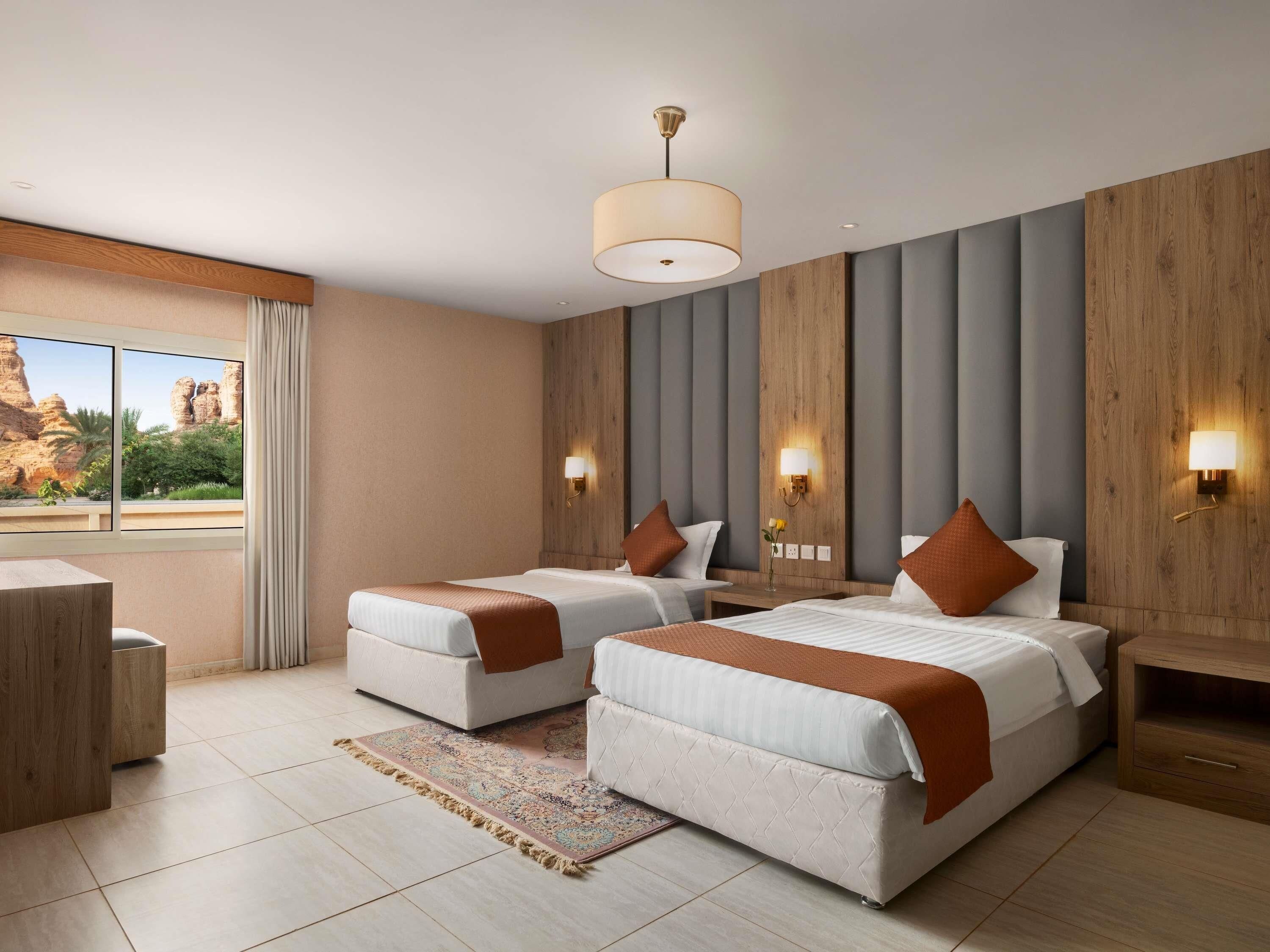 Quarto de hotel com duas camas, uma escrivaninha, uma janela grande com vista e um lustre moderno.