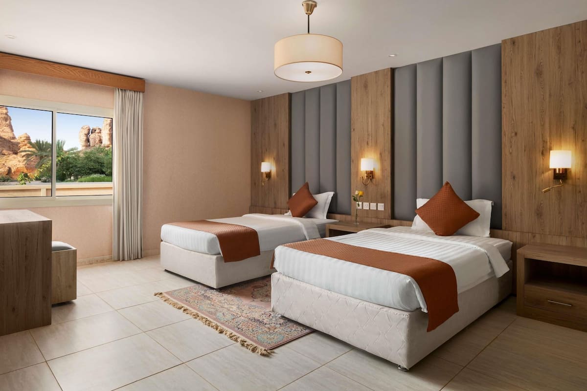 Quarto de hotel com duas camas, uma escrivaninha, uma janela grande com vista e um lustre moderno.
