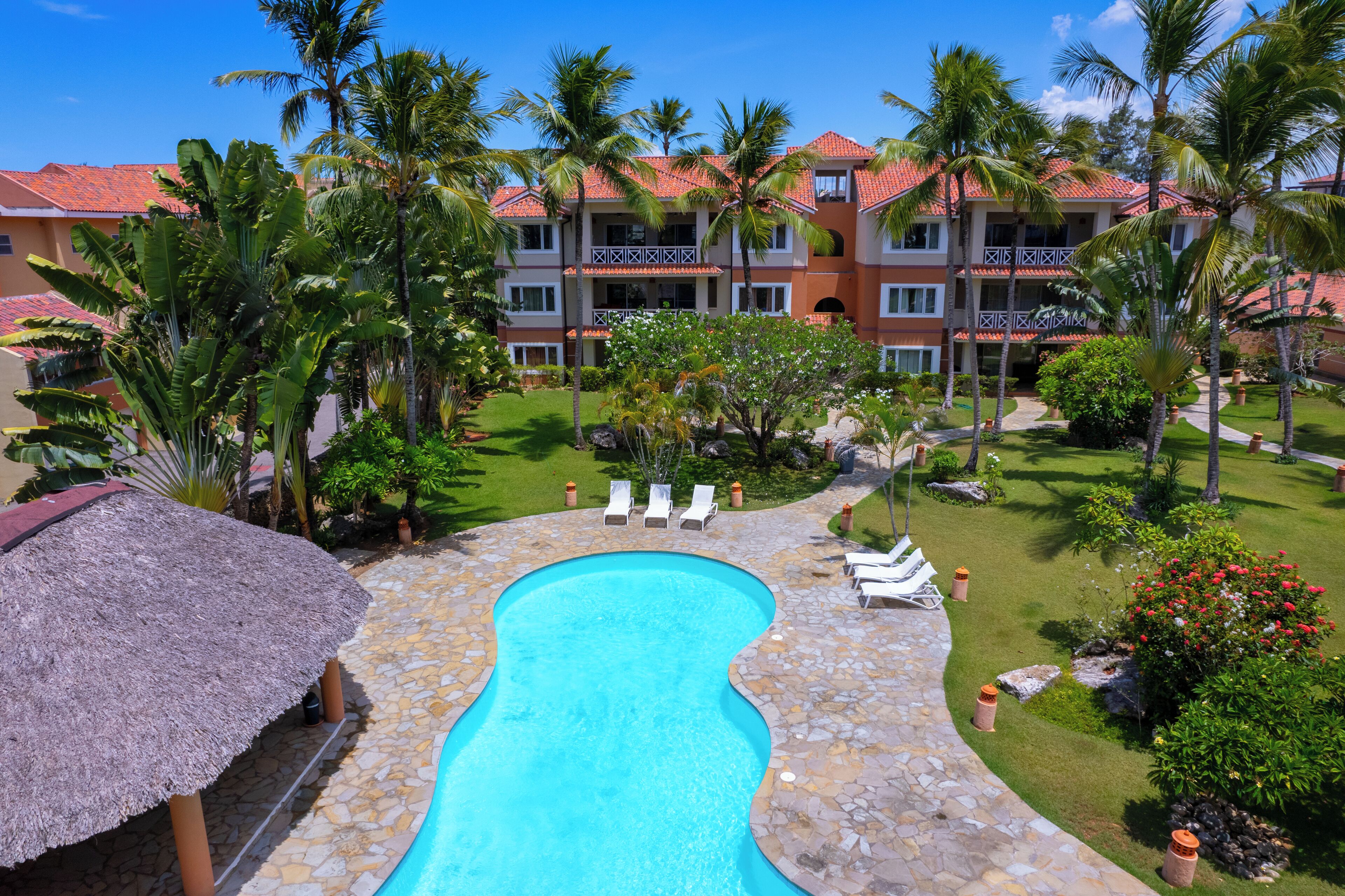 Piscina com espreguiçadeiras, cercada por palmeiras e um edifício de hotel com telhados de telhas vermelhas.