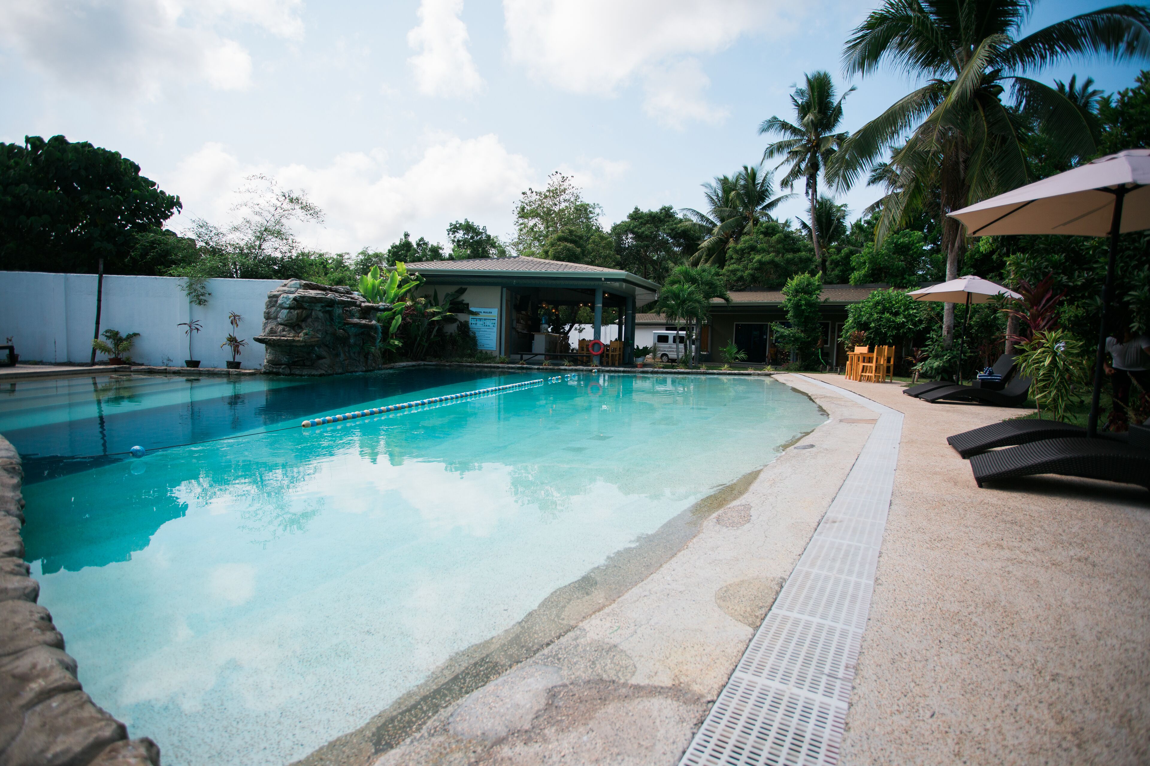Piscina com água azul cristalina, rodeada por espreguiçadeiras e guarda-sóis, e uma área coberta com telhado.