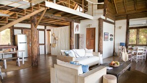 Um quarto espaçoso com piso de madeira, cama com roupa de cama branca, um banco de madeira, uma mesa de jantar com cadeiras e uma janela grande.