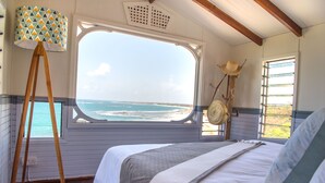Quarto com uma grande janela com vista para a praia, uma cama com roupa branca, piso de madeira e um abajur estampado.