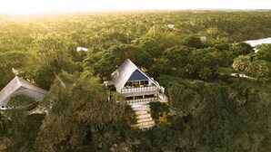 Uma casa rodeada por uma floresta densa, com um telhado triangular imponente e uma escadaria que leva à entrada.