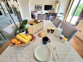 Uma área de refeições com uma mesa posta para dois, apresentando uma cesta de palha com frutas, um prato, talheres e potes.