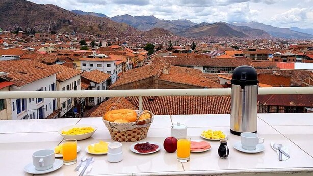 Uma mesa de café da manhã com diversos alimentos e uma garrafa térmica, com vista para uma paisagem urbana com edifícios de telhado vermelho e montanhas ao fundo.