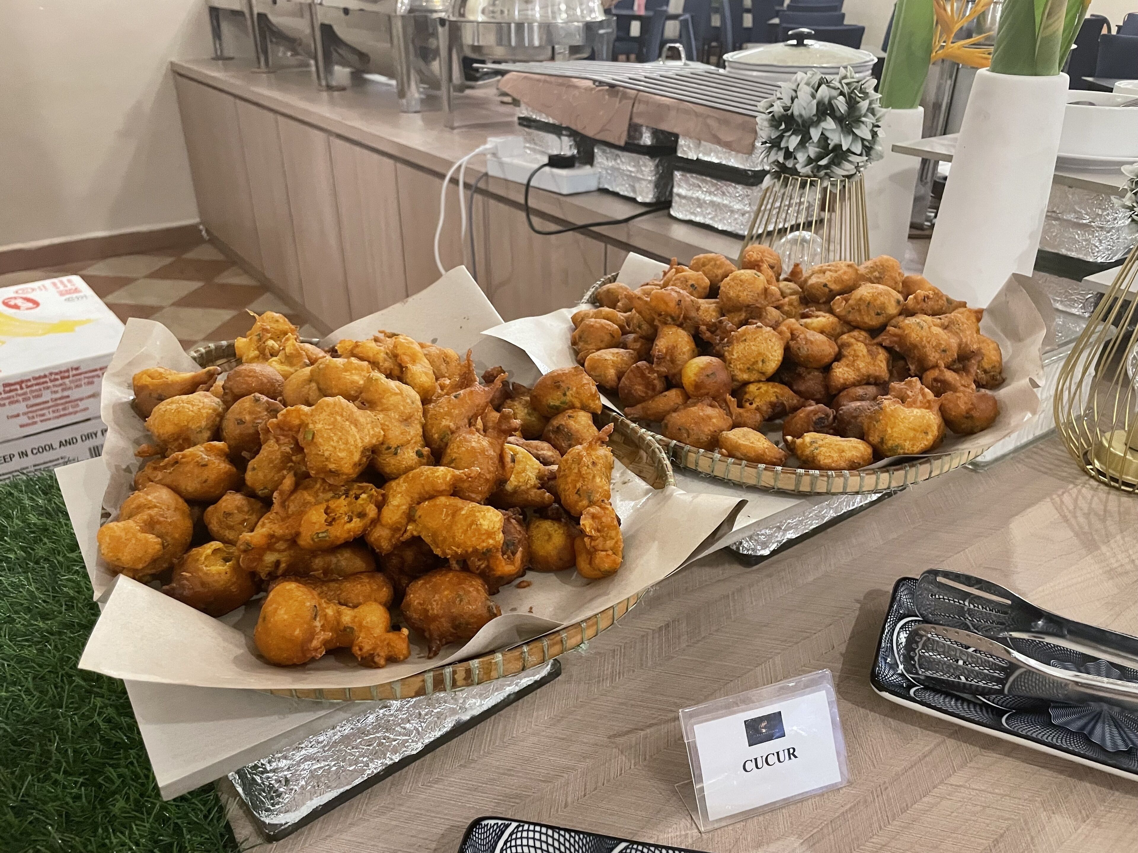Uma mesa de buffet com bandejas de alimentos fritos, uma placa com a inscrição 'CUCUR' e um par de pinças do lado direito.