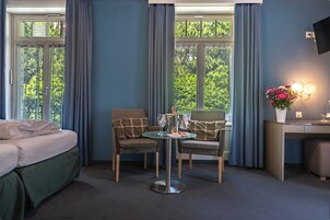 Quarto de hotel com uma cama, duas cadeiras, uma mesinha, uma escrivaninha e uma janela grande com cortinas.