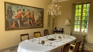 Uma sala de jantar com um grande quadro clássico, um candelabro e uma mesa posta para refeição.