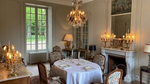 Uma sala de jantar com uma mesa redonda posta para seis pessoas, rodeada por cadeiras elaboradas. Uma janela grande com vista para o verde é visível, e uma lareira com um espelho e castiçais está ao fundo.
