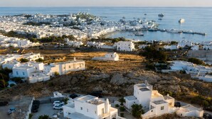 Vista aérea de uma estância costeira com edifícios brancos, uma piscina e barcos ao fundo.