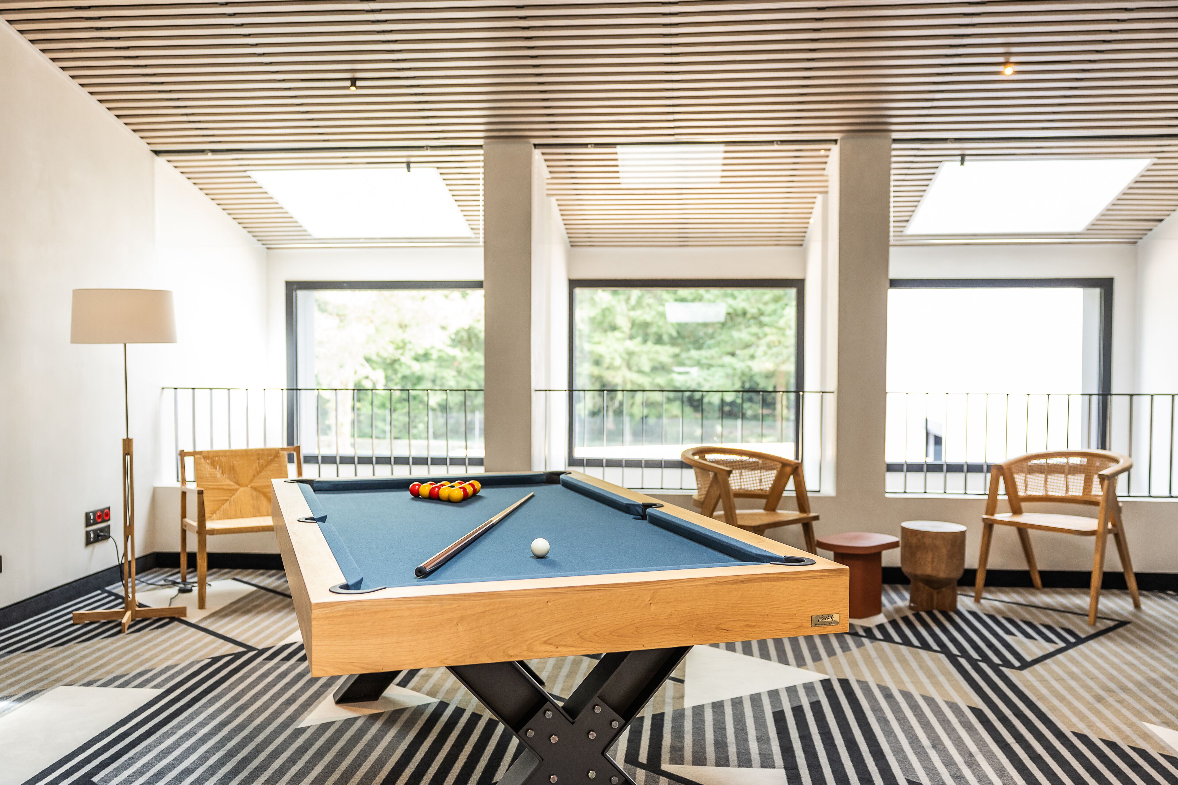 Uma mesa de bilhar em um espaço com janelas amplas, cadeiras de madeira e uma luminária.