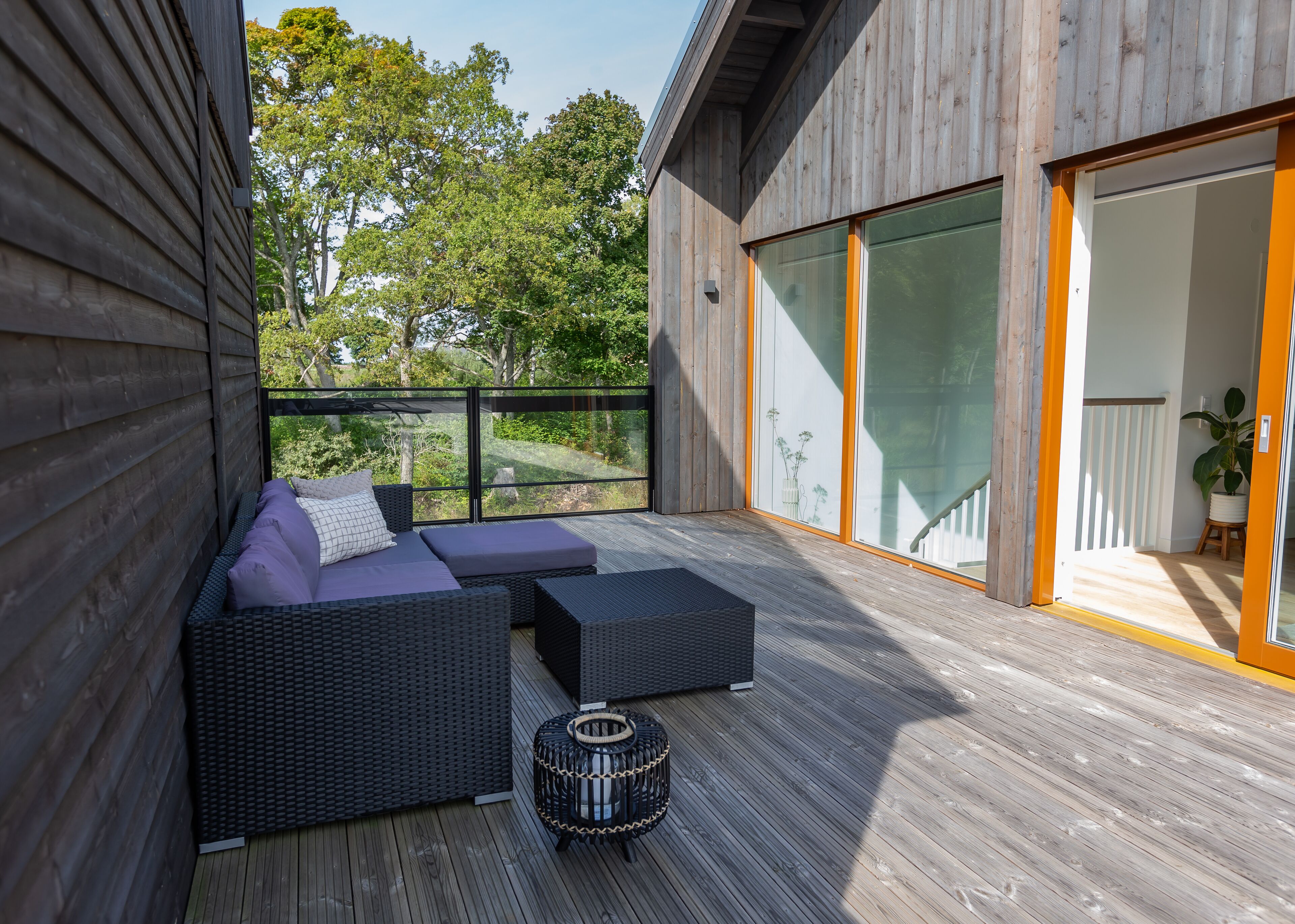 Um deck de madeira com um sofá e uma mesa pequena, uma porta de vidro que dá para uma varanda, e vista para árvores.