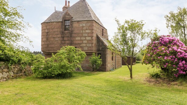 Um edifício de pedra com telhado inclinado, rodeado por um gramado e árvores.