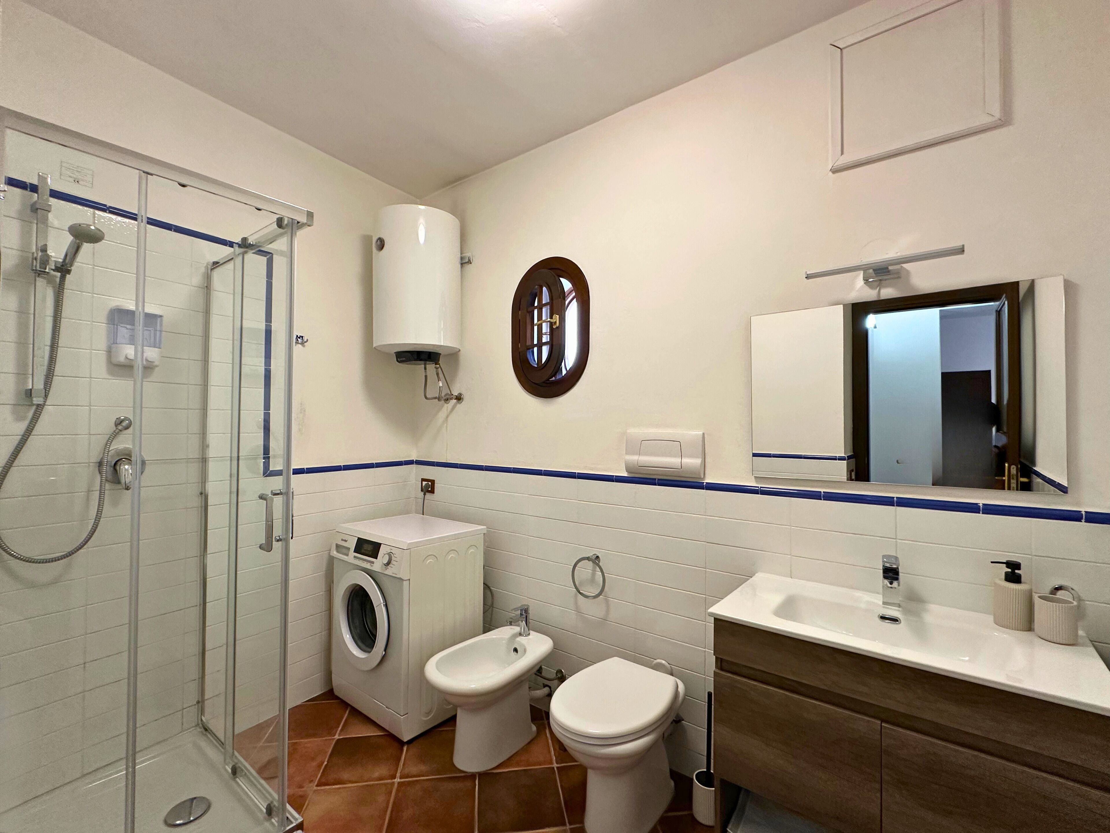 Casa de banho com chuveiro separado por vidro, máquina de lavar roupa, bidé, sanita e lavatório com espelho em cima.