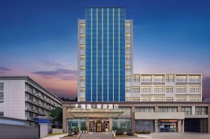 Um hotel com design moderno, apresentando um edifício central imponente com fachada de vidro, entrada térrea com área coberta e um prédio menor adjacente.