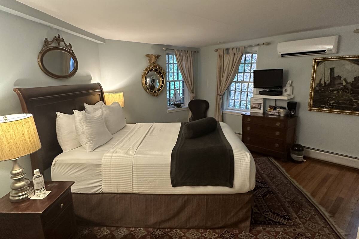 Um quarto bem arrumado com uma cama grande, uma mesa de cabeceira com abajur, cômoda e uma televisão sobre um suporte.