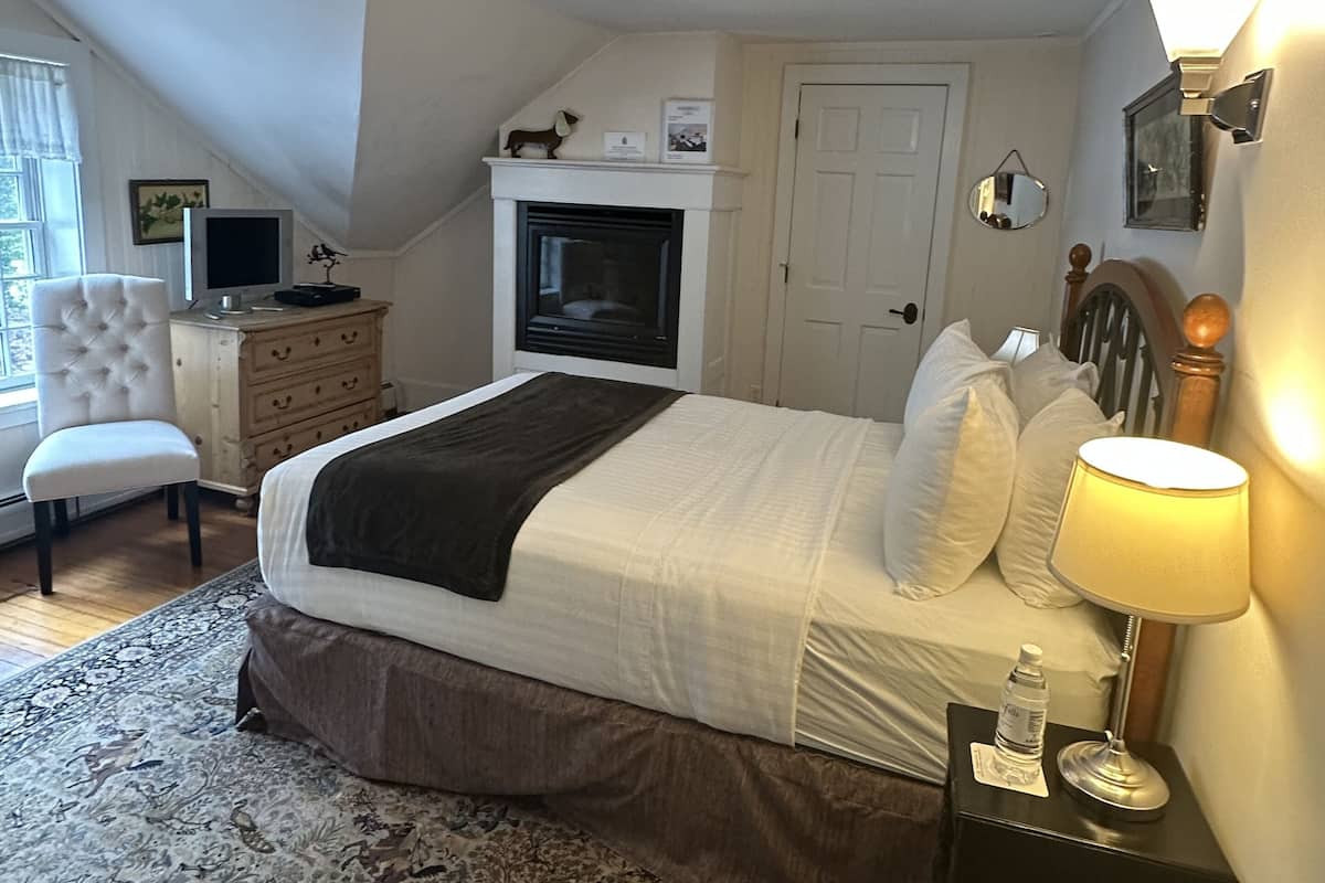 Quarto com cama, luminária de cabeceira, cômoda, cadeira e lareira.