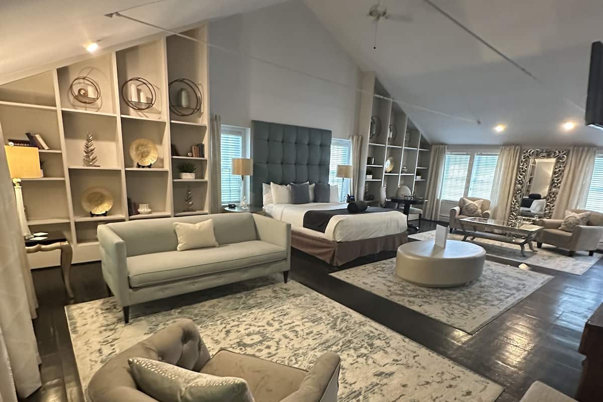 Uma sala de estar espaçosa com um sofá grande, um banquinho redondo e uma mesa de centro. Há prateleiras com objetos decorativos e uma cama com cabeceira estofada.