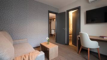 Quarto de hotel moderno com parede cinza estampada, sofá cinza, mesa lateral de madeira com um vaso, cadeira branca e televisão de tela plana na parede.