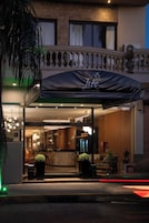 Entrada de hotel com área externa coberta, balcão de recepção e interior visível com assentos e plantas.
