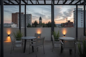 Um pátio externo com mesas e cadeiras redondas, plantas em vasos e vista para edifícios da cidade e um campanário ao entardecer.