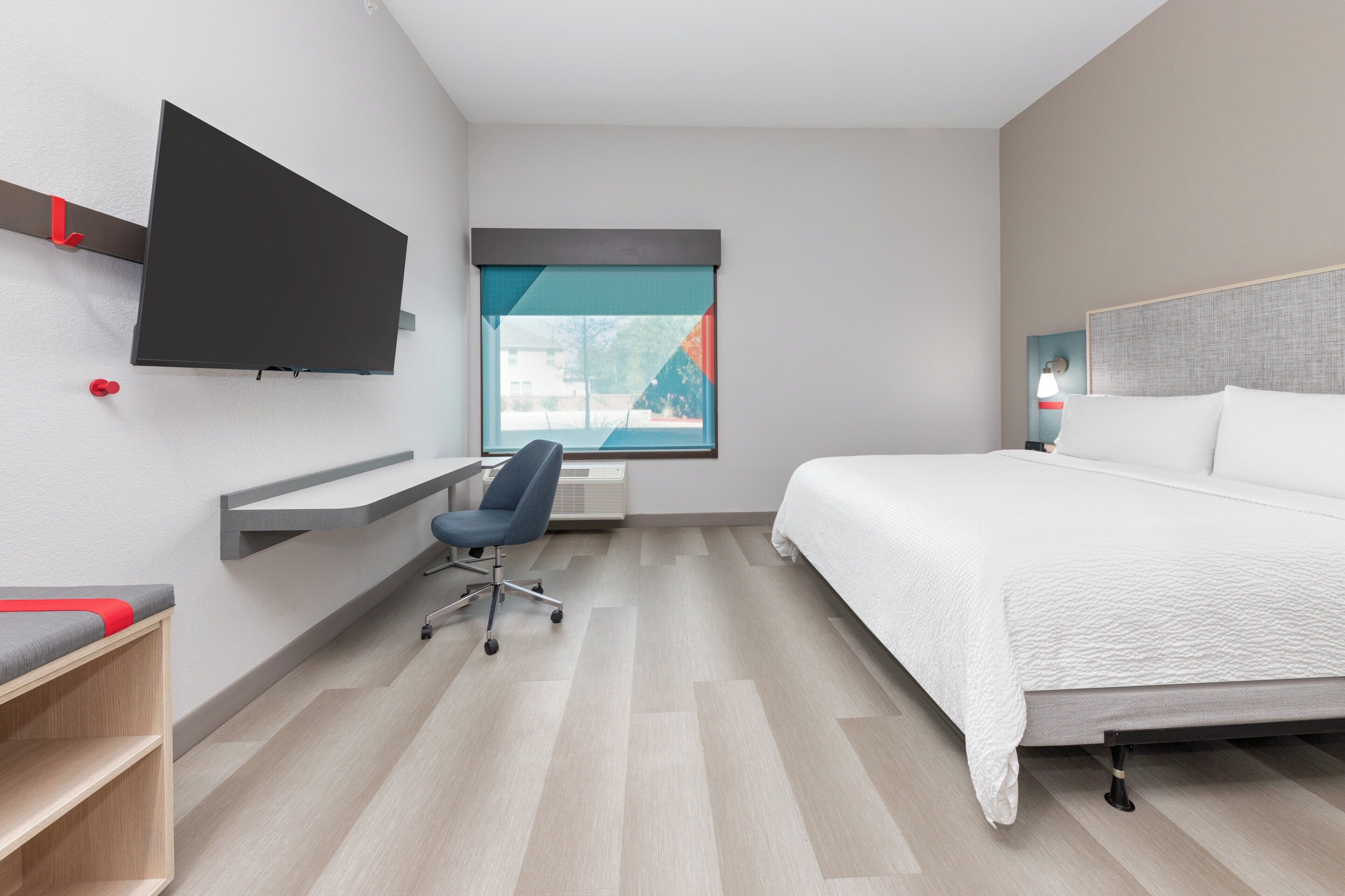 Quarto de hotel moderno com cama, escrivaninha, cadeira, TV fixada na parede e janela com vista para o exterior.