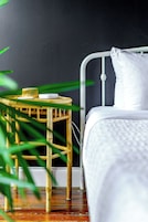 Uma cama com roupa de cama branca, uma mesa de cabeceira com um aparelho pequeno e um banquinho de bambu.