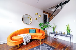 Uma sala de estar moderna com um sofá laranja curvo, um espelho redondo, uma mesa de console com plantas e uma escada.