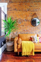 Quarto com parede de tijolos, um espelho redondo, sofá de couro marrom, manta de tricô amarela e uma planta verde num cesto trançado.