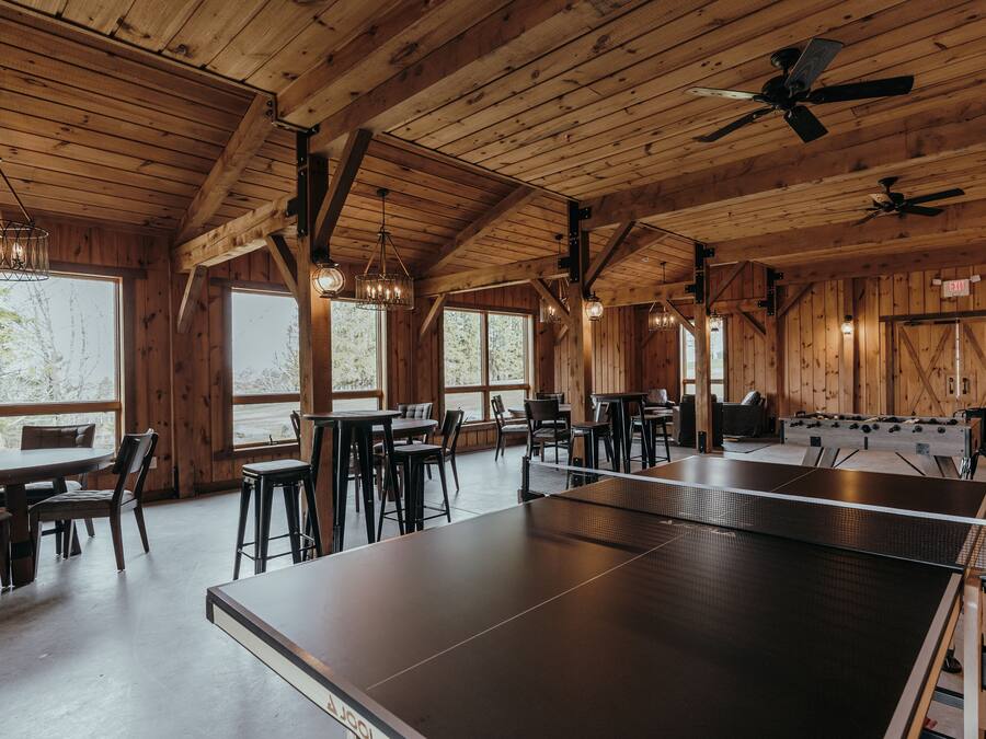 Interior de madeira com mesa de tênis, mesas, cadeiras e grandes janelas.