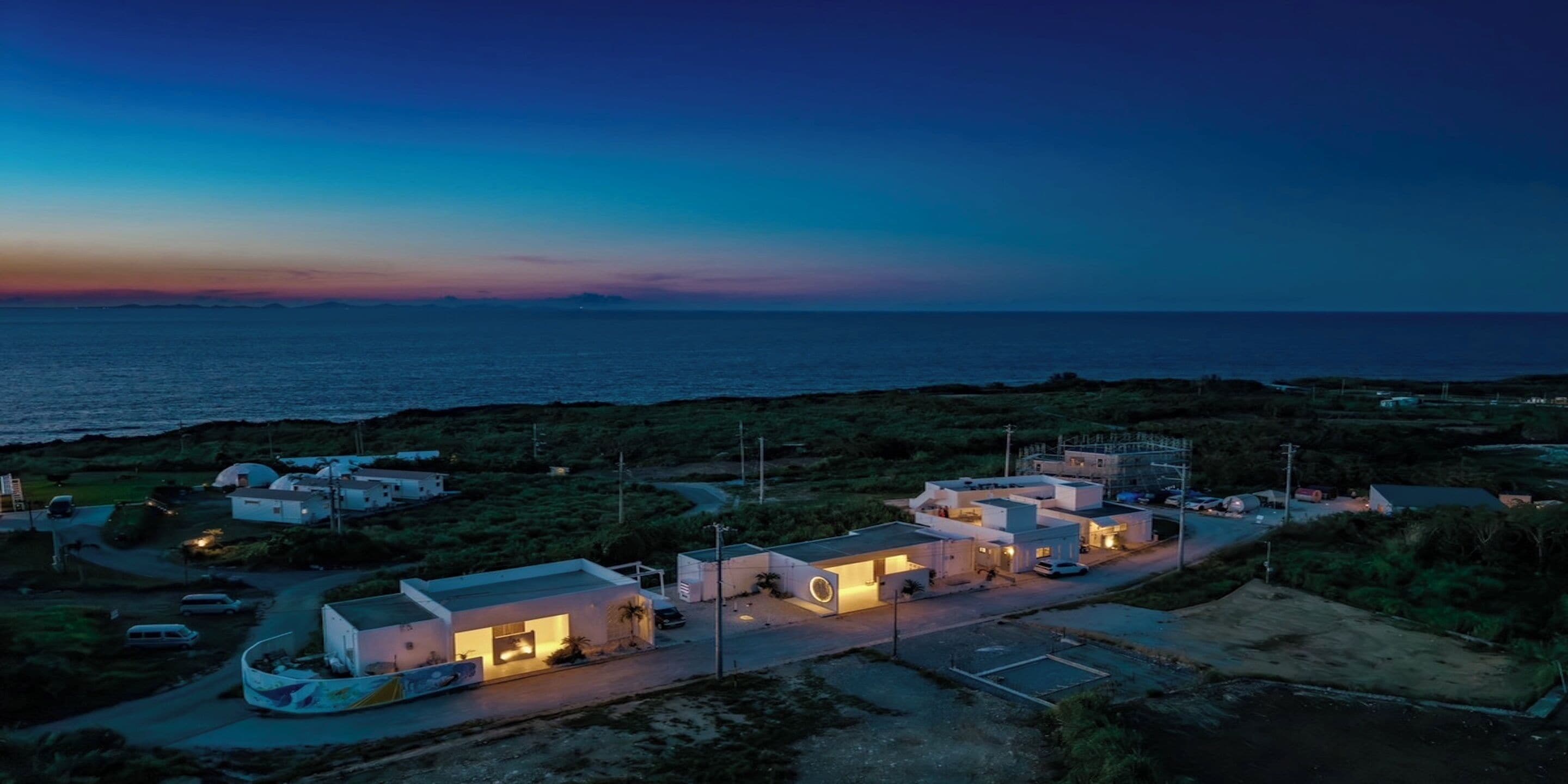 Zona residencial costeira com várias casas, uma estrada e vista para o mar ao entardecer.