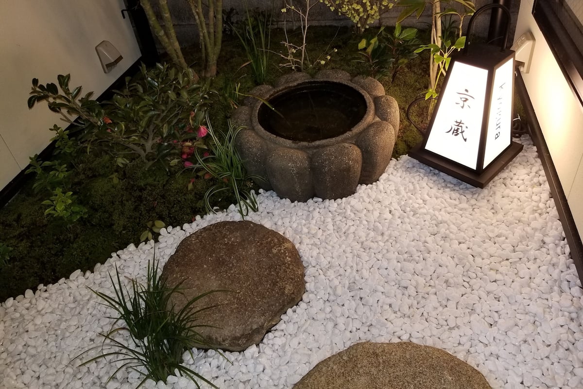 Um jardim com uma bacia de pedra, pedrinhas brancas e diversas plantas.