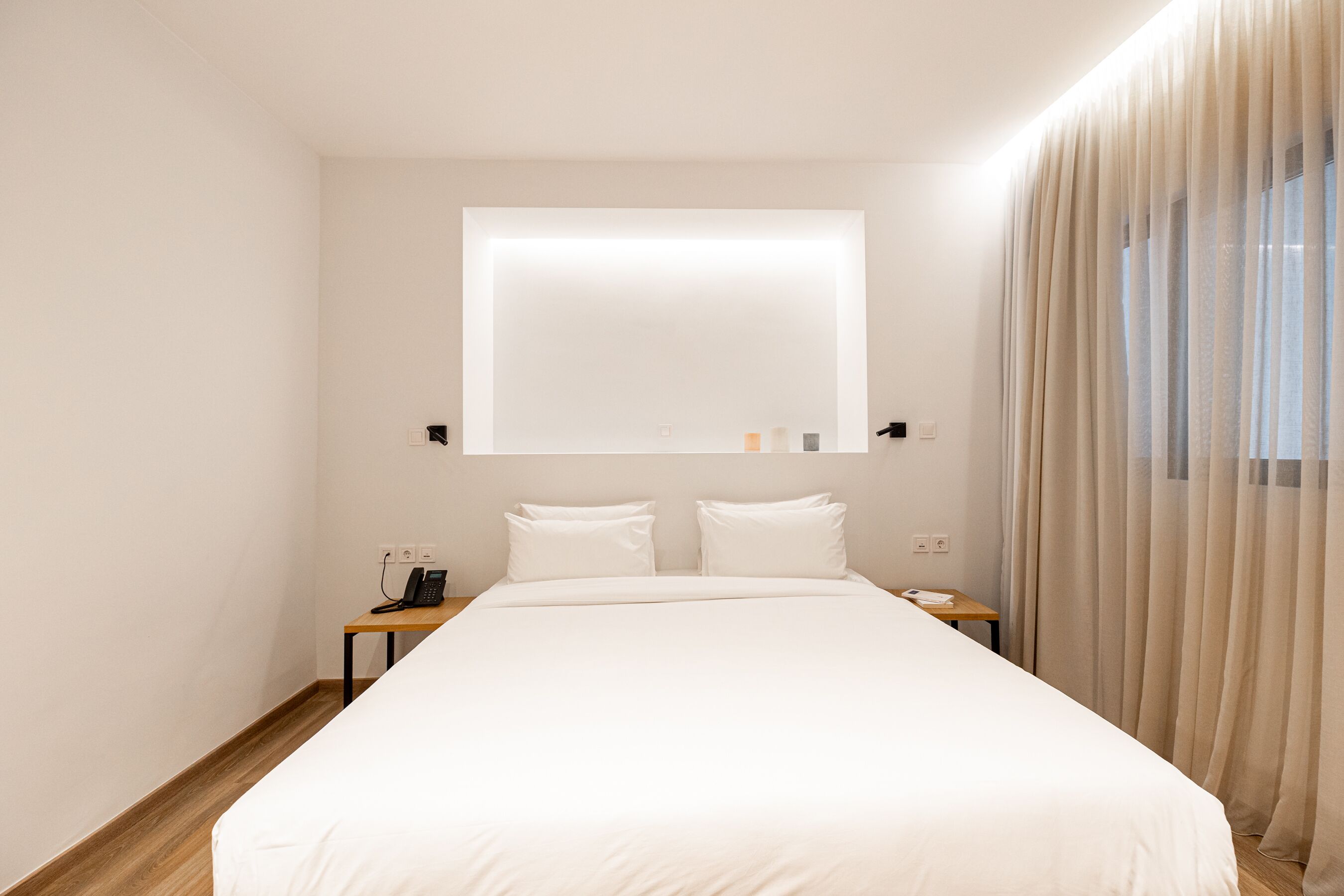 Quarto de hotel moderno com uma cama grande, mesas de cabeceira, luz acoplada à parede e vista através de cortinas leves.