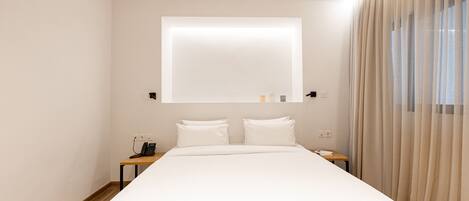 Quarto de hotel moderno com uma cama grande, mesas de cabeceira, luz acoplada à parede e vista através de cortinas leves.