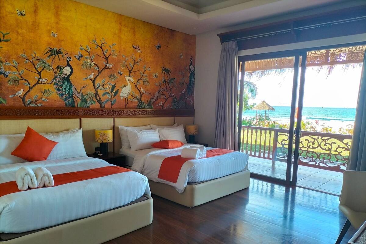 Quarto de hotel com duas camas, um mural grande na parede e uma varanda com vista para a praia.