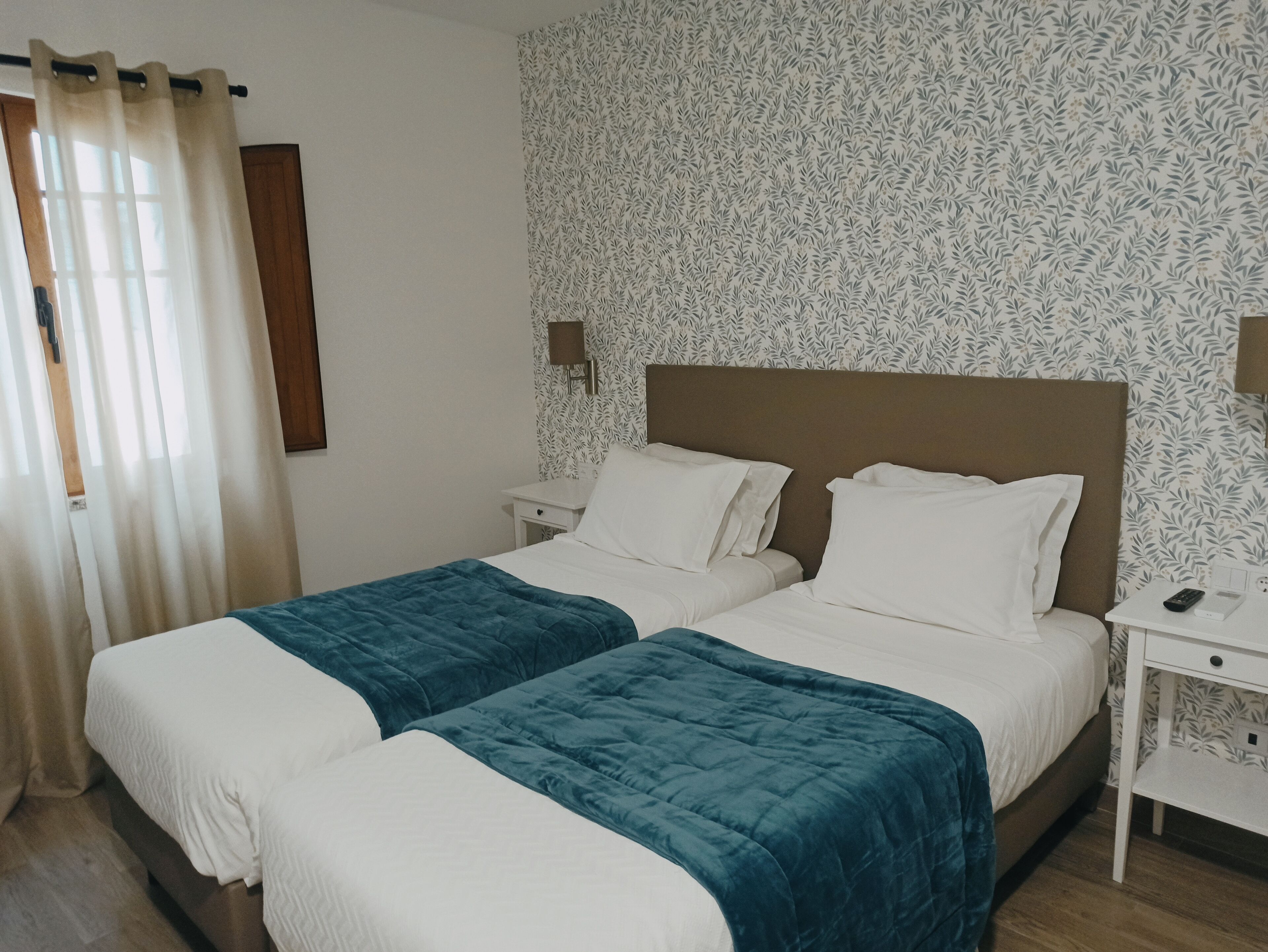 Uma cama de casal com cabeceira marrom, roupa de cama branca e manta verde-água. Há uma mesinha de cabeceira branca com uma luminária, uma porta de madeira e um papel de parede estampado.