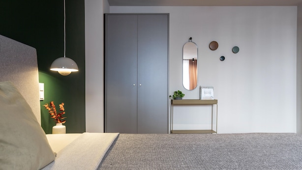 Quarto moderno com cama, mesa de cabeceira com espelho, luminária pendente e parede verde.