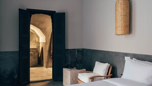 Quarto com uma cama, uma cadeira, uma mesinha de cabeceira, um cesto de palha e uma porta aberta que dá para o banheiro.