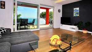 Uma sala de estar moderna com sofá, uma mesa de centro de vidro com frutas, TV e varanda com vista para o mar.