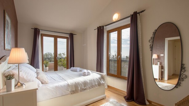 Quarto com uma cama grande, uma mesa de cabeceira com abajur, um espelho e vista para o exterior pelas janelas.