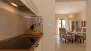 Cozinha moderna com fogão por indução embutido, área de refeições com cadeiras de vime e uma grande janela que permite entrada de luz natural.