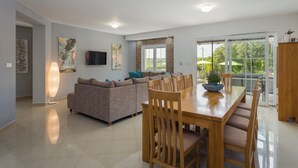 Uma sala de estar moderna com sofá, mesa de jantar e cadeiras. Há uma TV fixada na parede e uma grande janela que proporciona luz natural.