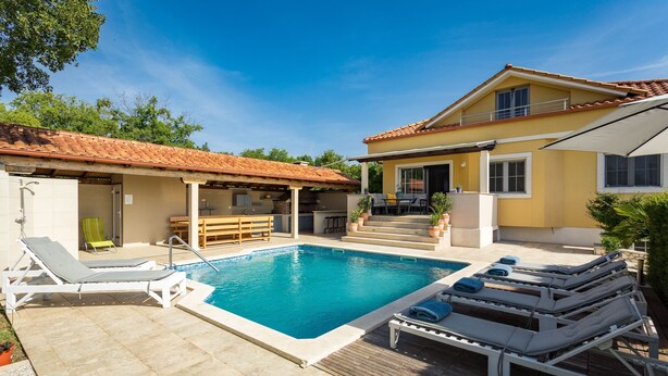 Uma piscina com espreguiçadeiras, um alpendre coberto e um edifício amarelo com telhado em azulejos.