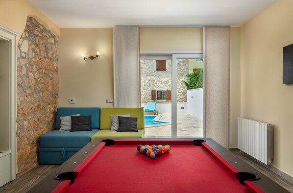Mesa de bilhar em um espaço com parede de pedra, sofá verde e vista para área externa com piscina.