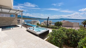 Uma área de piscina moderna ao ar livre, com hidromassagem, um terraço de pedra e vista para o mar e montanhas.