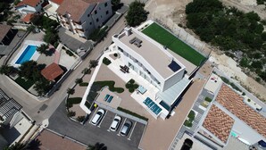Vista aérea de um edifício moderno com piscina no terraço, quadra de tênis e área de estacionamento.