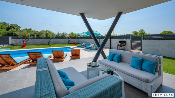 Uma área externa moderna para relaxar, com piscina, espreguiçadeiras e um sofá com almofadas azuis.